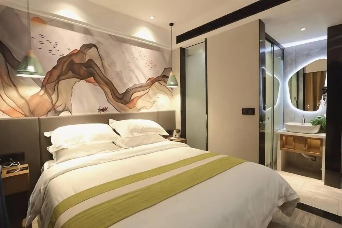 Quarto de hotel moderno com uma cama grande, uma obra de arte na parede, um banheiro com espelho e pia, e um edredom verde listrado.