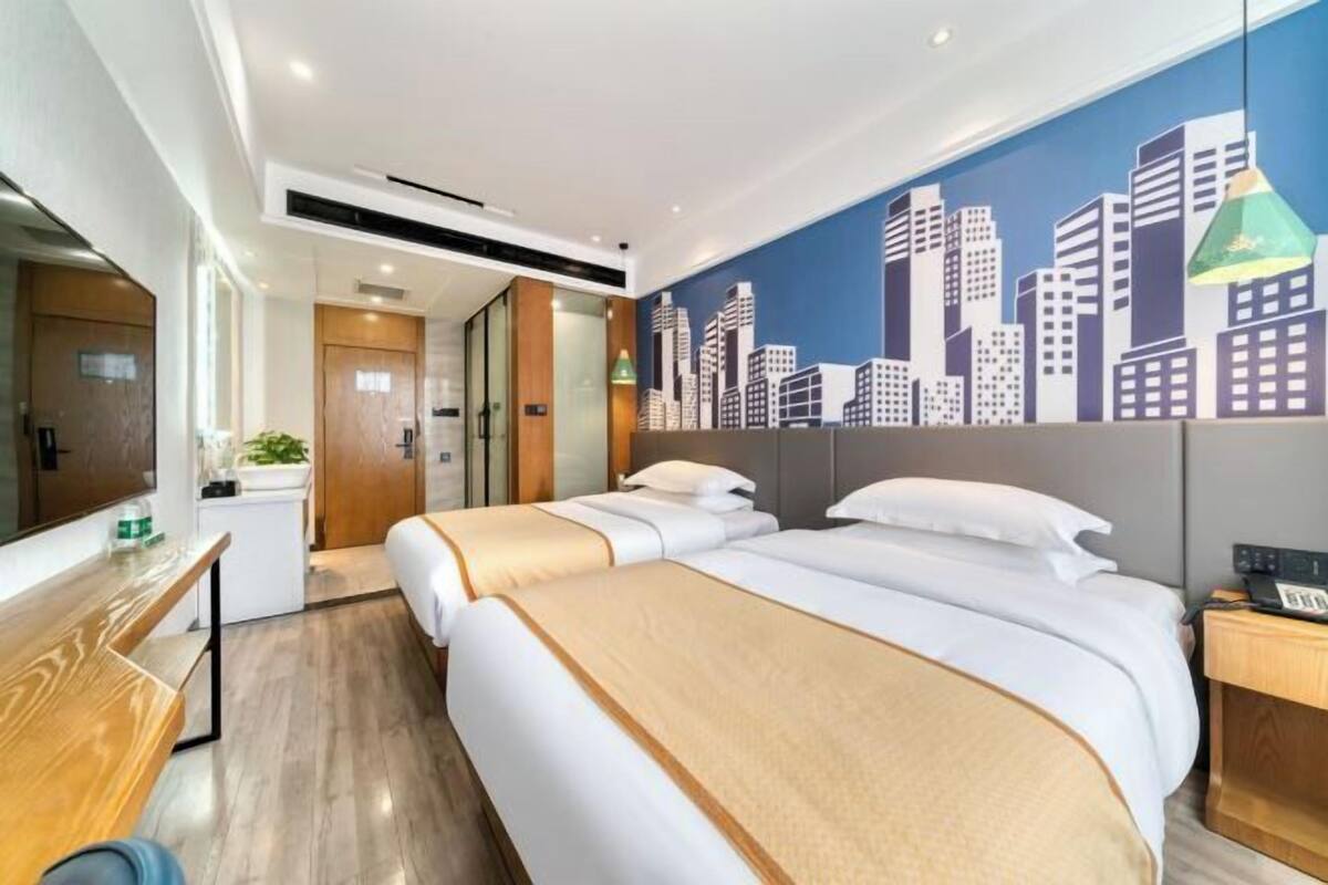 Quarto de hotel com um mural da paisagem urbana, uma cama com colcha bege, uma escrivaninha com cadeira e uma pequena cozinha.