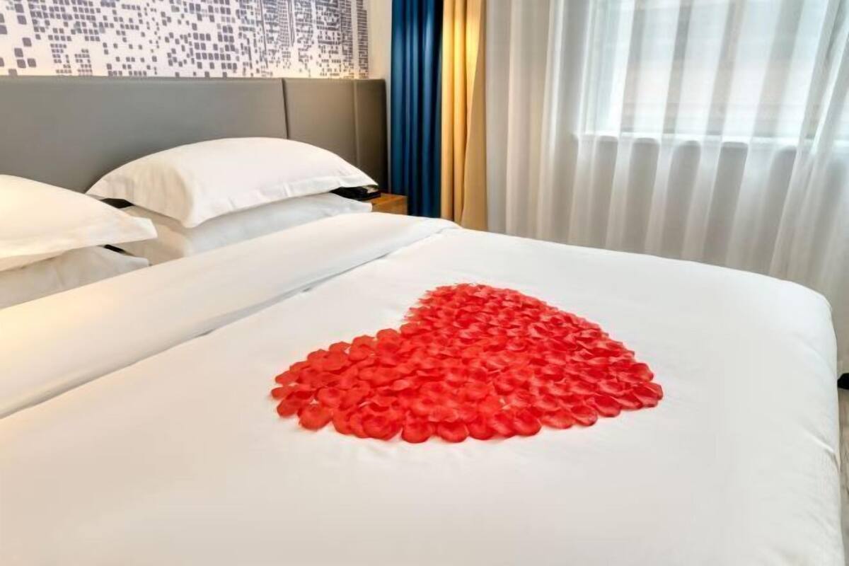 Quarto de hotel com cama arrumada, roupa de cama branca e um arranjo de pétalas de rosa vermelhas em formato de coração na cama.