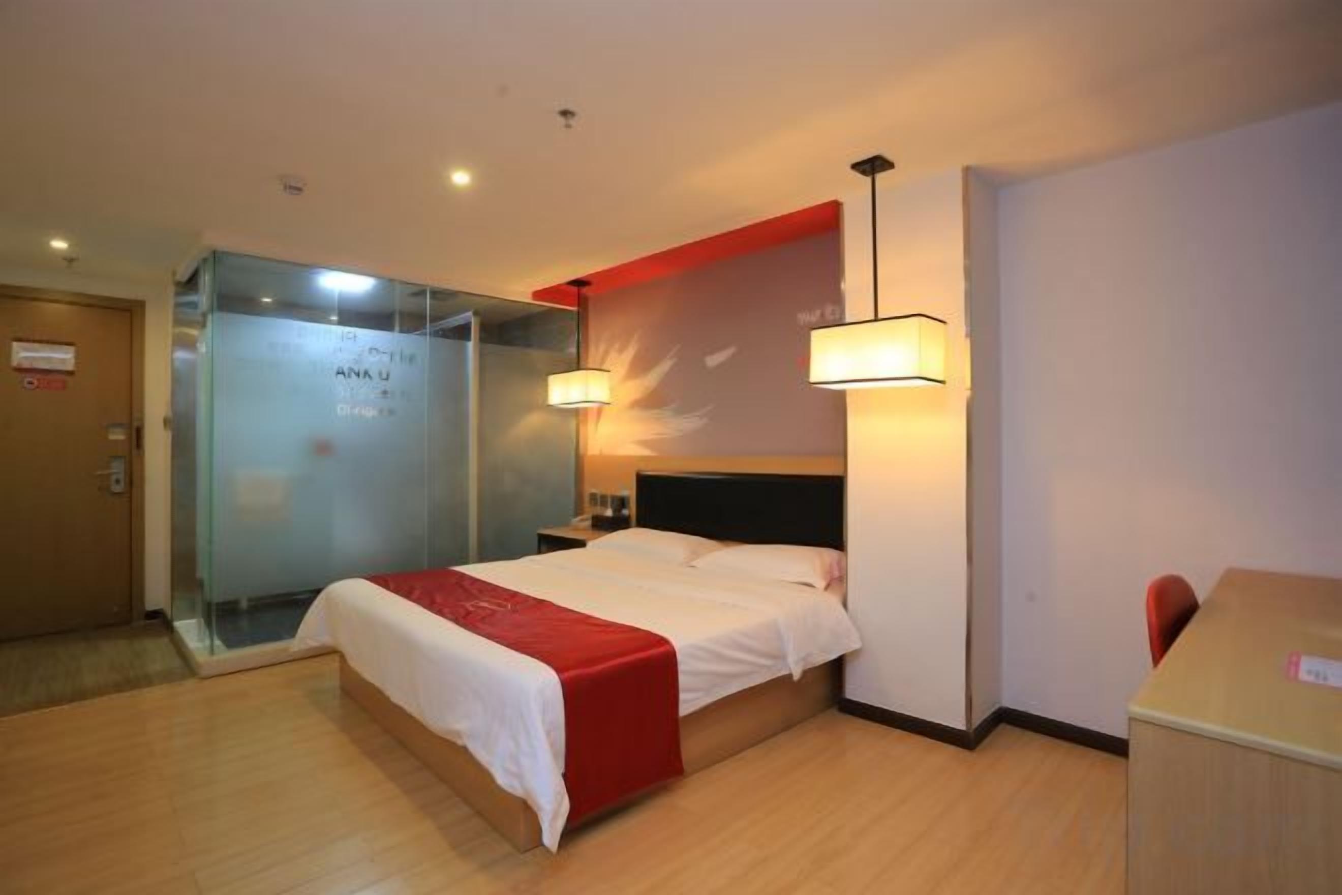 Quarto de hotel com cama, mesas de cabeceira, escrivaninha, box de vidro e luminária na parede.