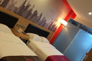 Quarto de hotel com duas camas, uma mesa de cabeceira, um abajur e uma parede com um mural da paisagem urbana.