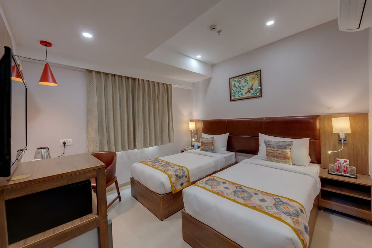 Quarto de hotel com duas camas, uma escrivaninha, uma cadeira, uma televisão, um abajur e uma janela com cortinas.