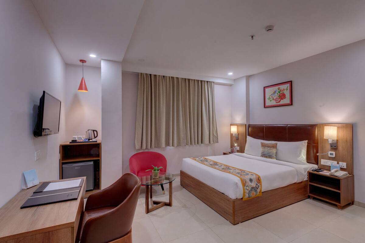 Quarto de hotel com cama, escrivaninha, cadeira, pequena mesa com planta, televisão, mini-geladeira, uma cadeira vermelha, mesa de cabeceira e luminária na parede.