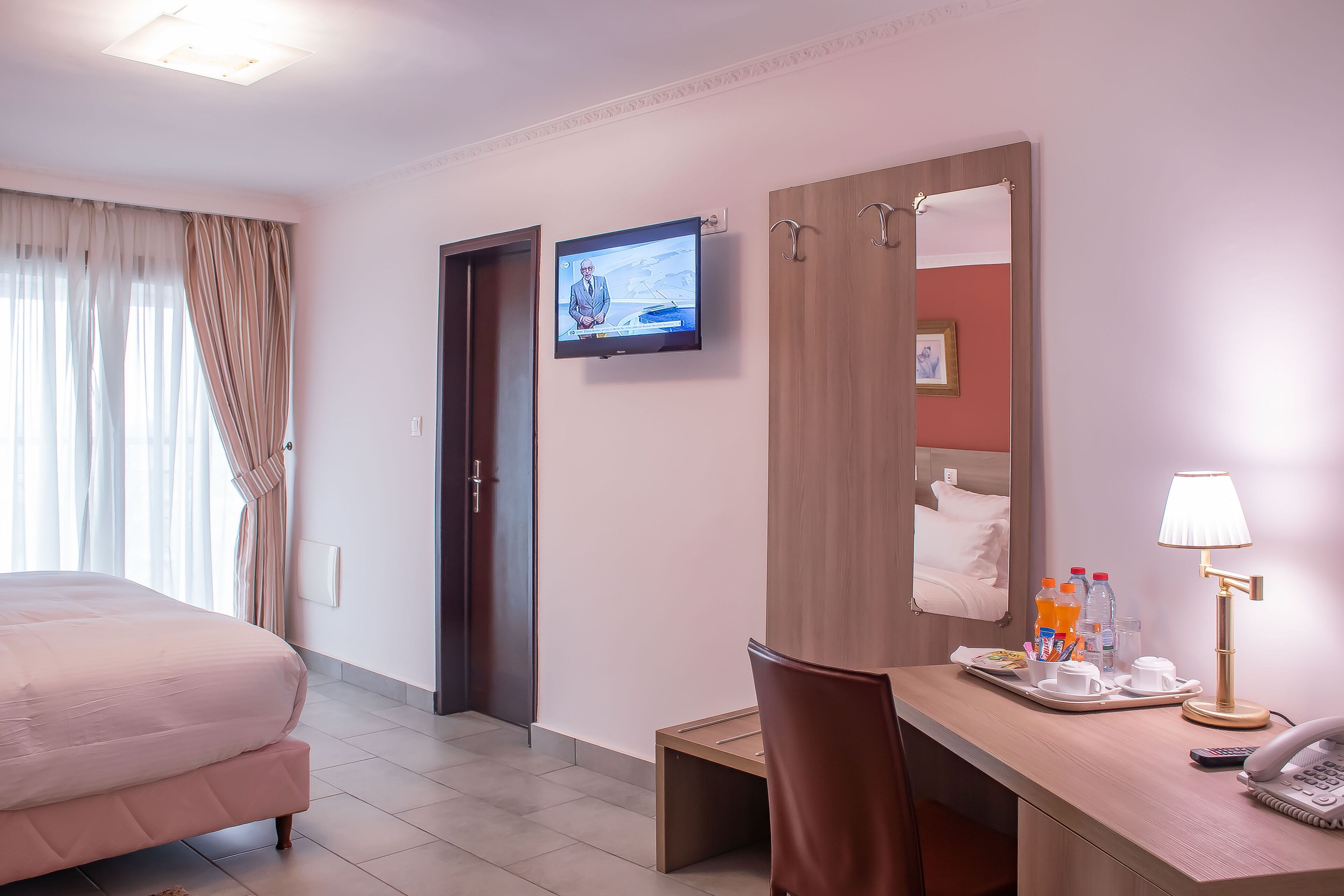 Quarto de hotel com cama, escrivaninha com luminária, televisão e espelho.