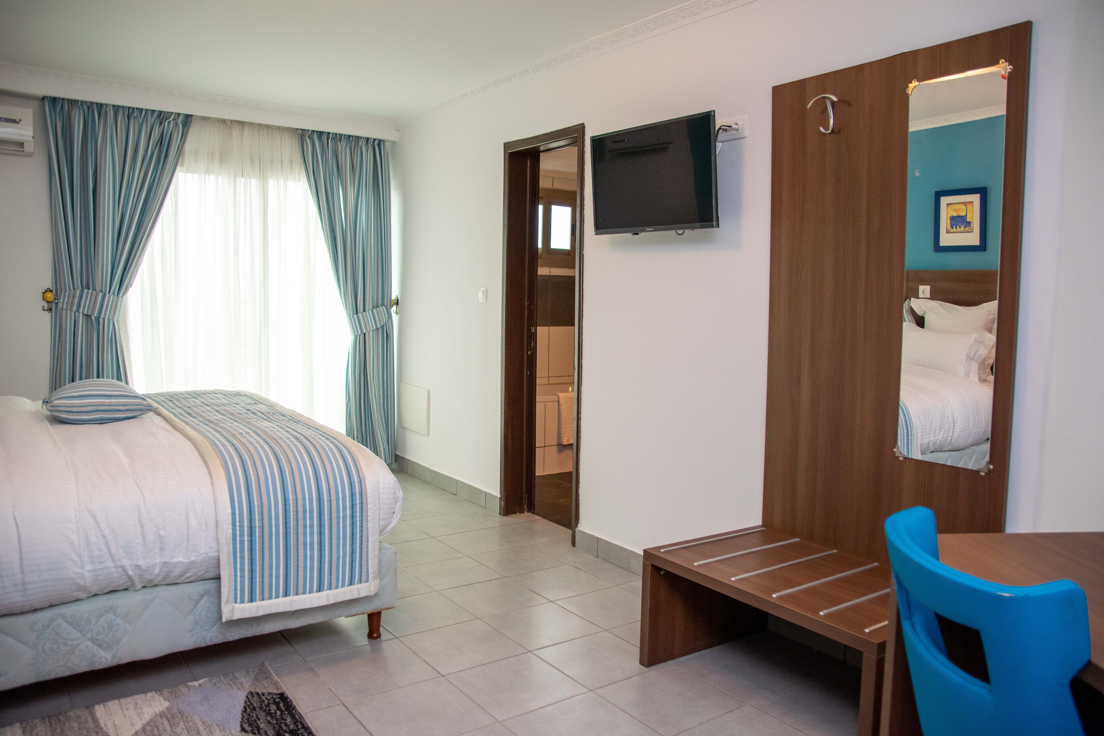 Quarto de hotel com cama, escrivaninha, cadeira, televisão e guarda-roupa.
