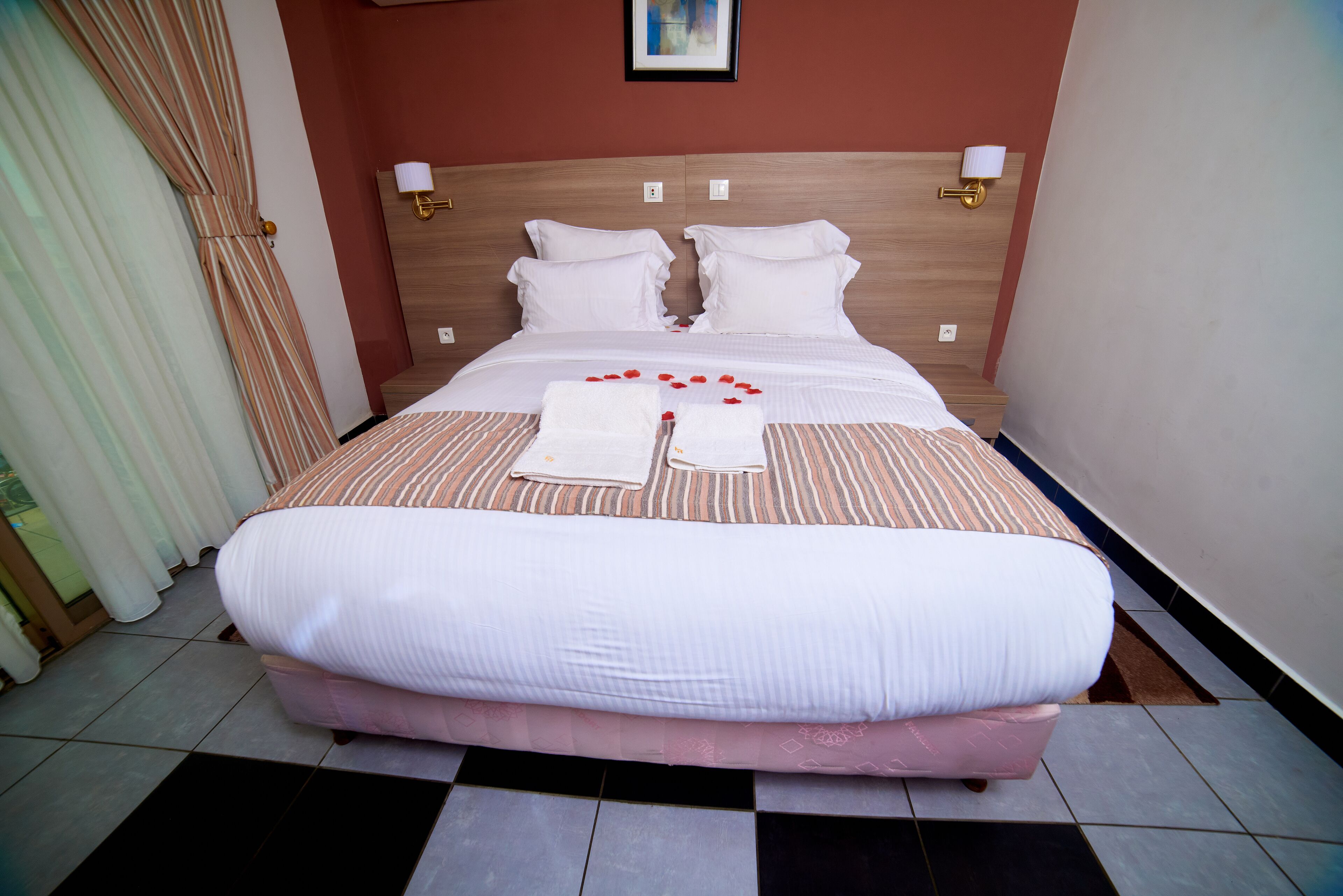 Cama bem arrumada com roupa de cama branca e listrada, cabeceira com luminárias fixadas na parede e um pequeno quadro acima da cabeceira.