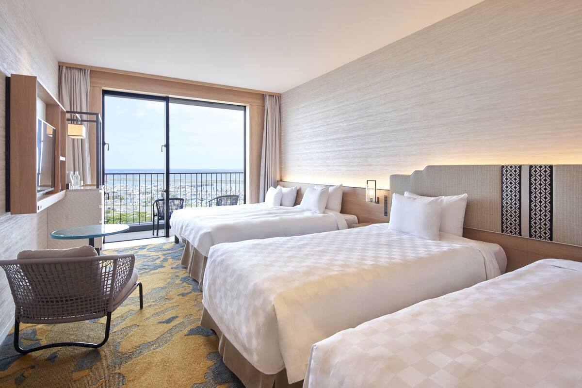 Quarto de hotel com duas camas, uma mesinha, uma cadeira e varanda com vista para a cidade.