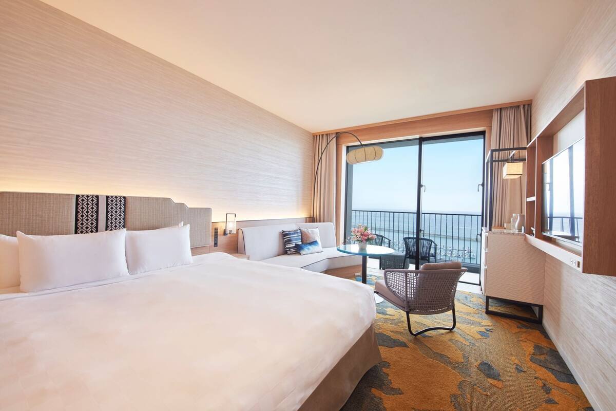 Quarto de hotel com uma cama grande, um sofá, uma mesa de jantar com cadeiras e uma varanda com vista.