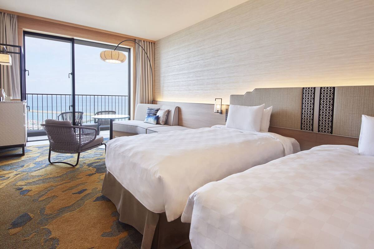 Quarto de hotel com duas camas, uma varanda com vista, uma escrivaninha e uma cadeira.