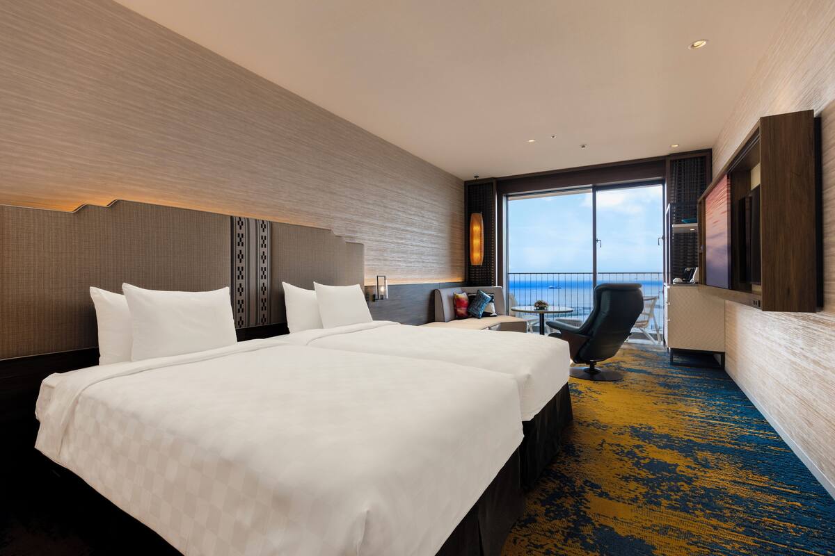 Quarto de hotel com uma cama grande, área de estar com uma cadeira, uma mesa pequena e uma janela ampla com vista.