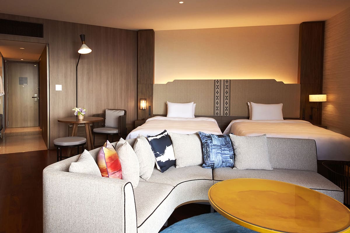 Quarto de hotel com sofá, duas camas, uma mesinha e uma luminária.