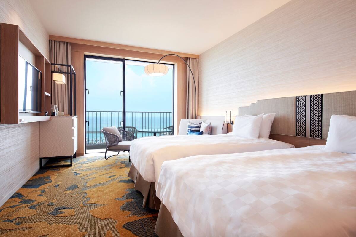 Quarto de hotel com duas camas, uma escrivaninha com televisão e varanda com vista.