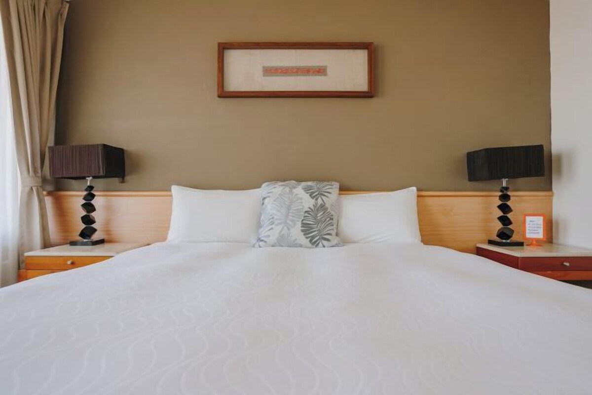 Uma cama bem arrumada com edredom branco, duas mesas de cabeceira com luminárias, um quadro na parede e cabeceira de madeira.