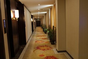 Um corredor de hotel com piso revestido de carpete, luminárias na parede e plantas em vasos.