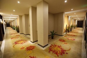 Corredor de hotel com piso coberto por carpete, colunas e plantas em vasos.