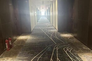 Um corredor comprido e estreito, com piso estampado, extintores de incêndio à esquerda e portas em ambos os lados.