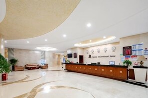 Uma área de recepção de hotel com piso de mármore, balcão de atendimento, espaço de espera com sofás e uma grande planta.