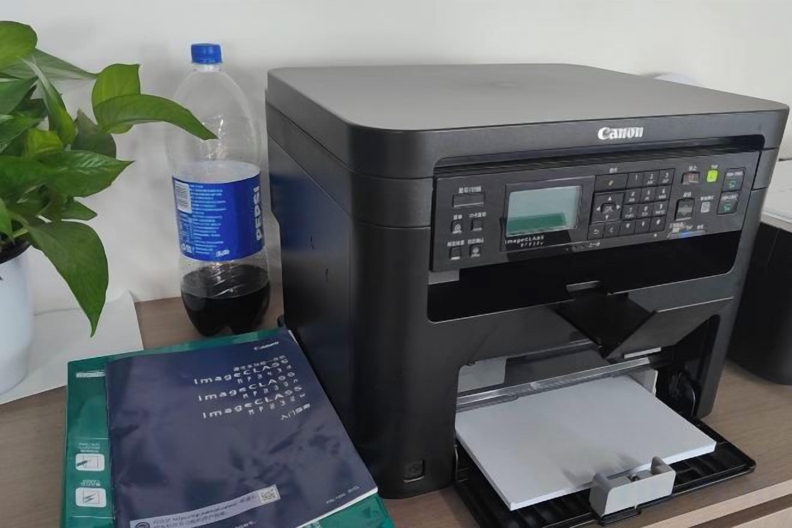 Uma impressora Canon sobre uma mesa, com uma garrafa de plástico, um saco de tinta para impressora e um vaso de planta.