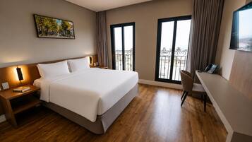 Quarto de hotel com uma cama grande, uma escrivaninha, uma cadeira, um quadro na parede e uma varanda com vista.