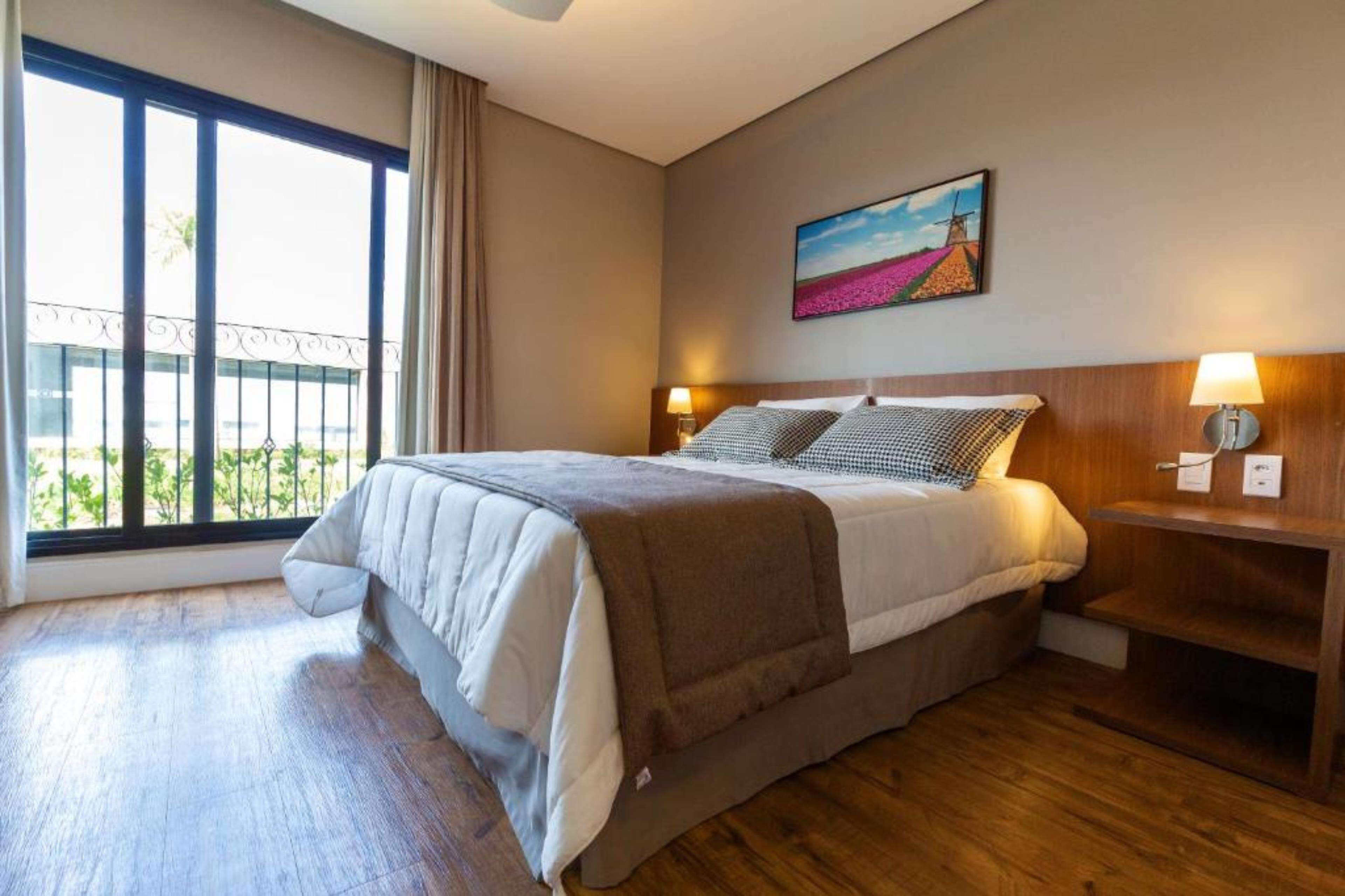 Quarto de hotel com uma cama grande, janela com vista, um quadro na parede e uma mesa de cabeceira com abajur.