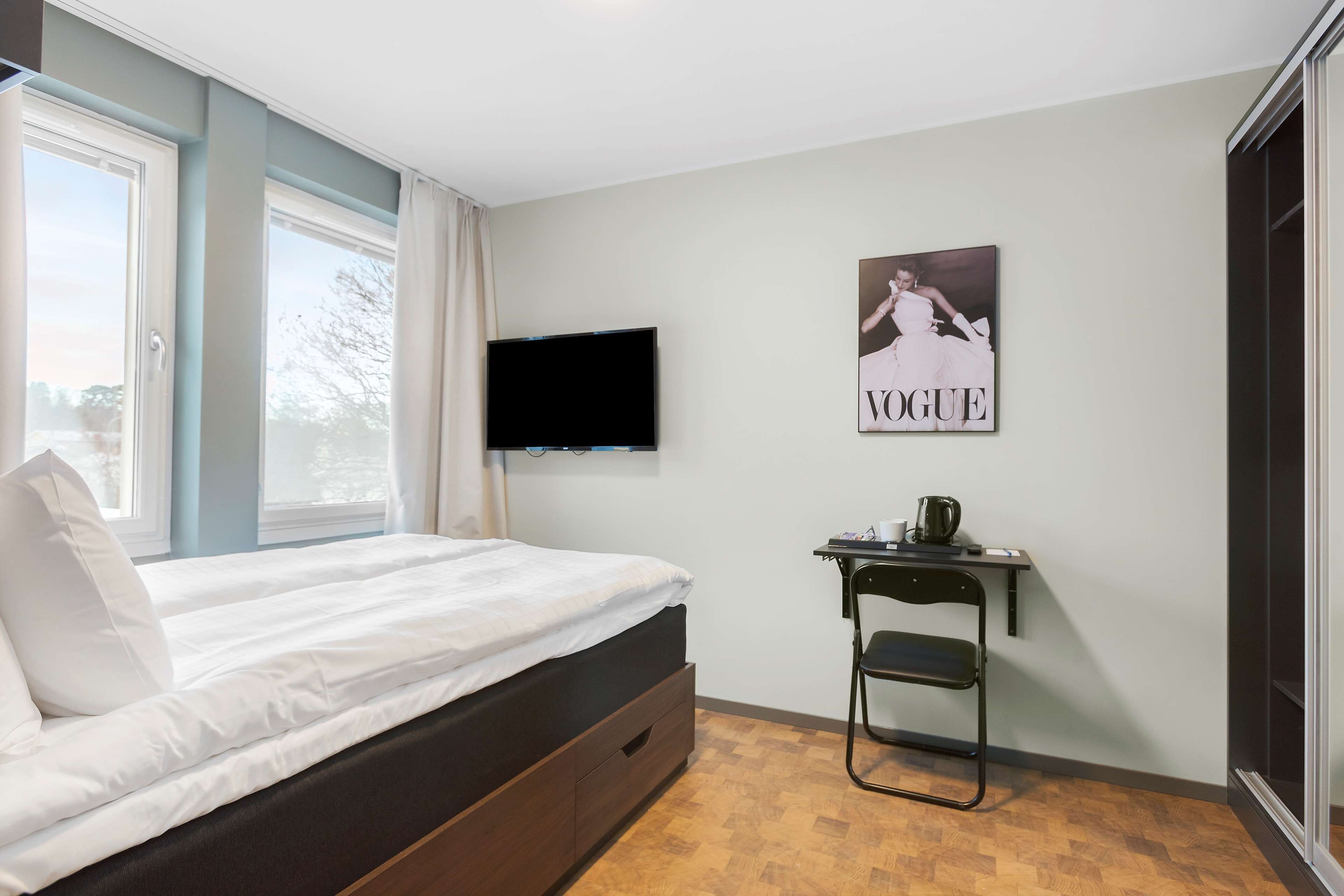 Quarto com uma cama, TV fixada na parede, pequena mesa com cadeira e um pôster na parede.