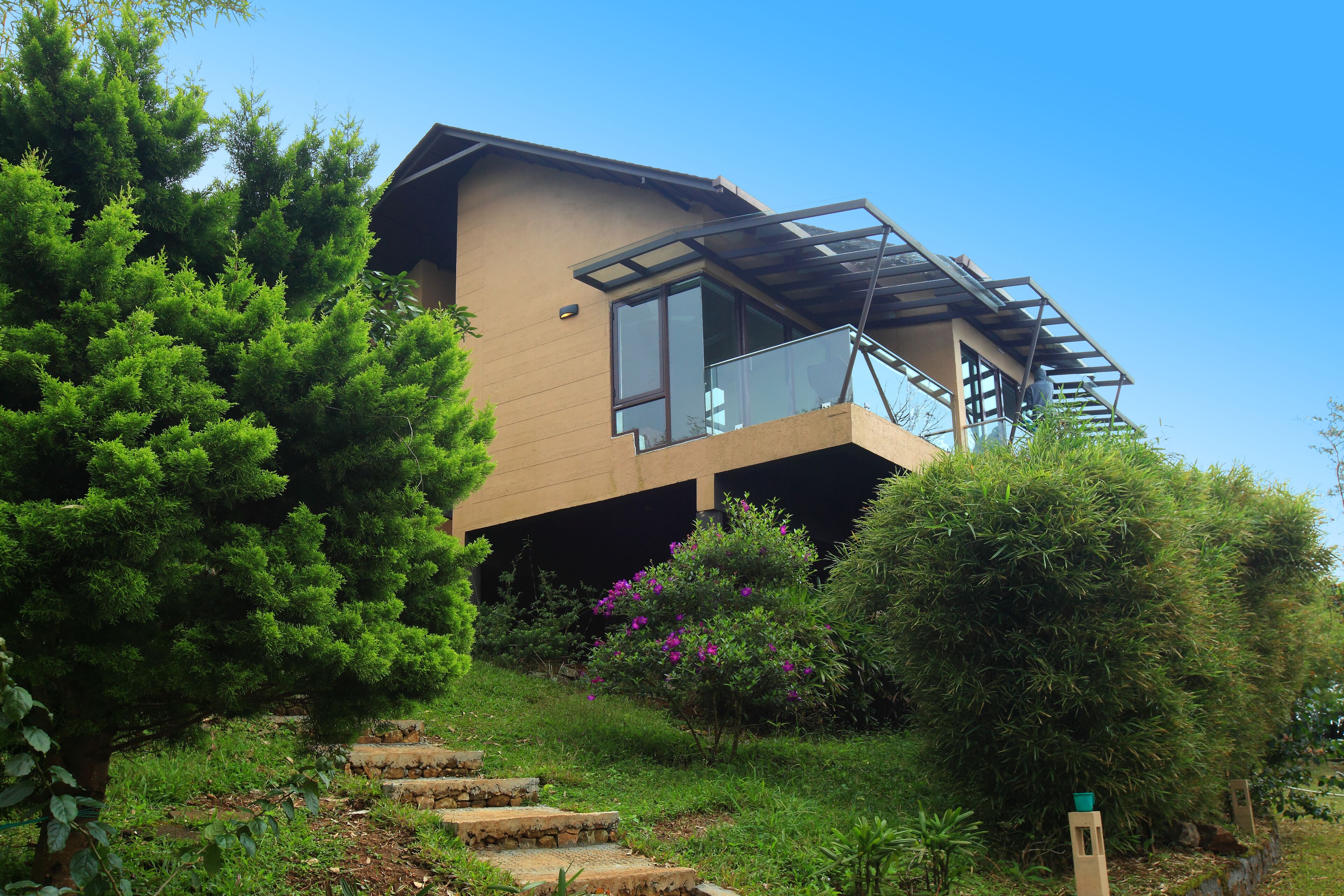 Uma casa moderna com varanda de vidro, rodeada por vegetação exuberante e uma escadaria de pedra.