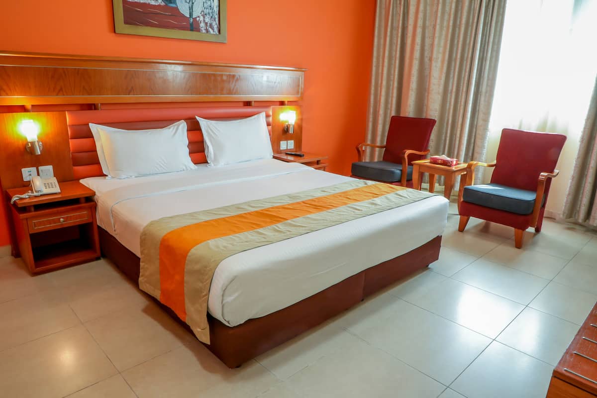 Quarto de hotel com uma cama grande, duas mesas de cabeceira com abajures, uma cabeceira de madeira e duas cadeiras vermelhas.