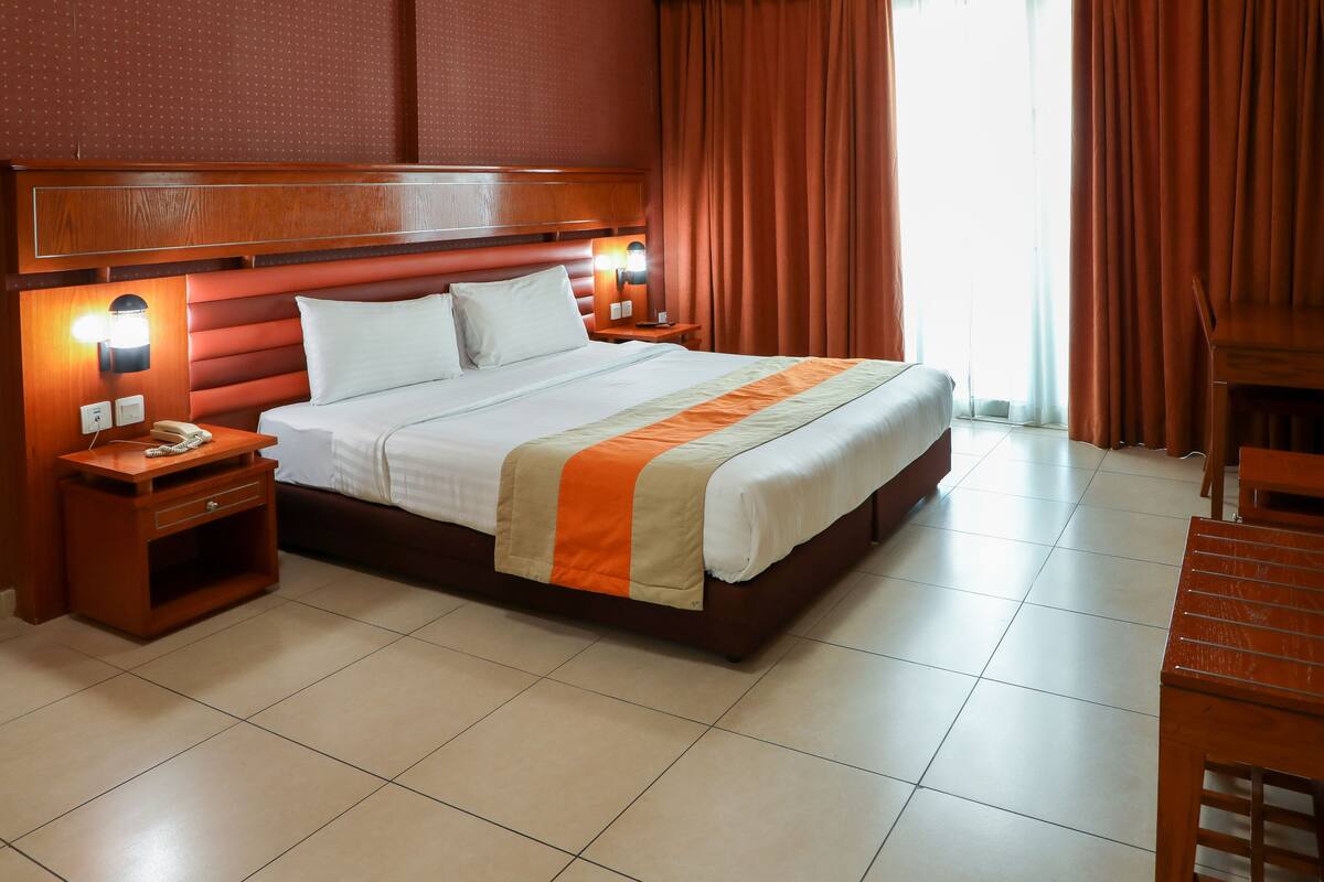 Quarto de hotel com uma cama grande, cabeceira de madeira, duas mesas de cabeceira com luminárias, uma mesinha de madeira e uma escrivaninha com cadeira.
