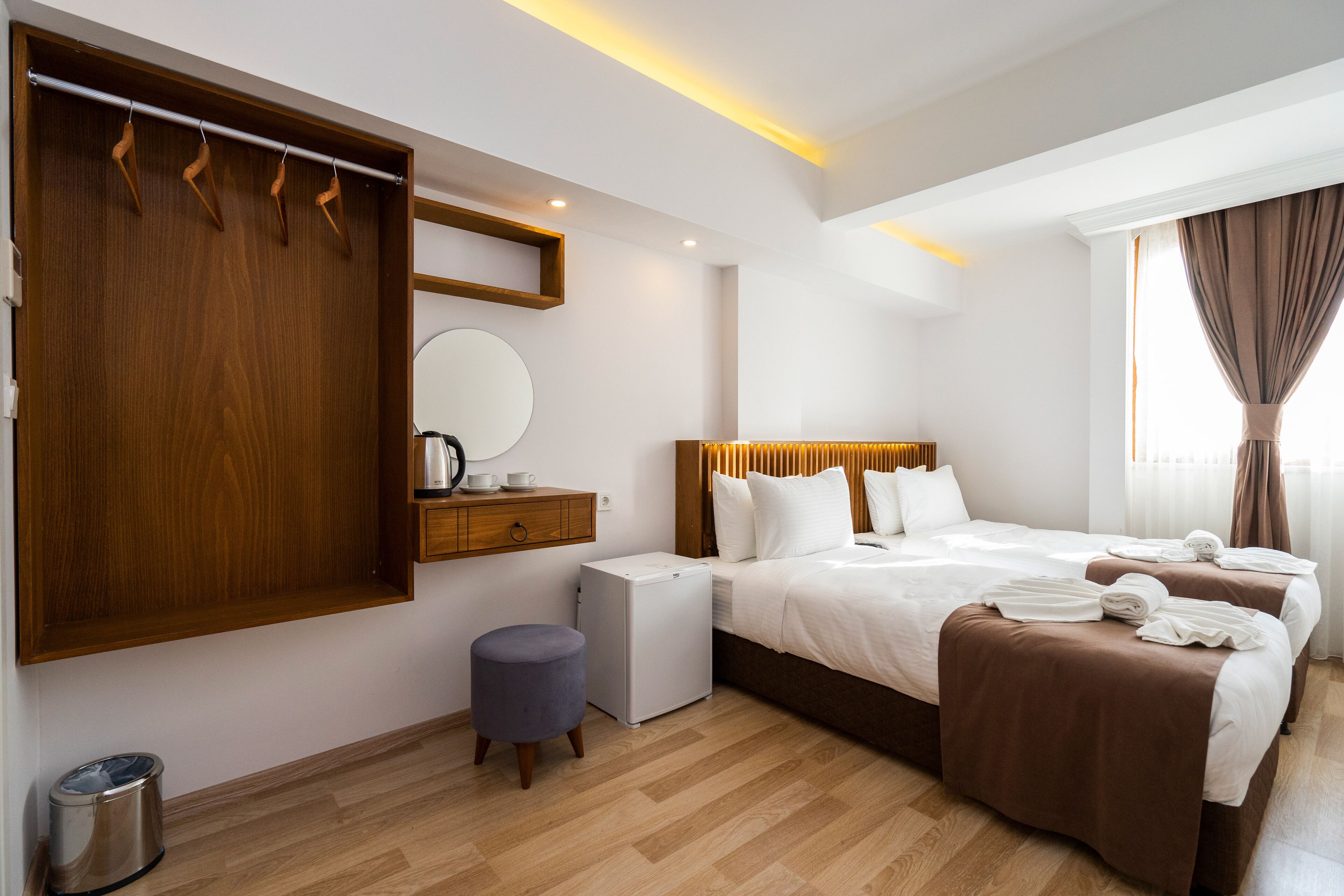 Quarto de hotel moderno com guarda-roupa de madeira, mesa de cabeceira pequena com chaleira, mini-frigorífico, espelho redondo e duas camas com roupa de cama castanha.