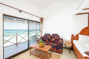 Quarto com cama de madeira, um sofá estampado com motivos tropicais, uma mesa com frutas e vista para o oceano através de uma porta de correr.
