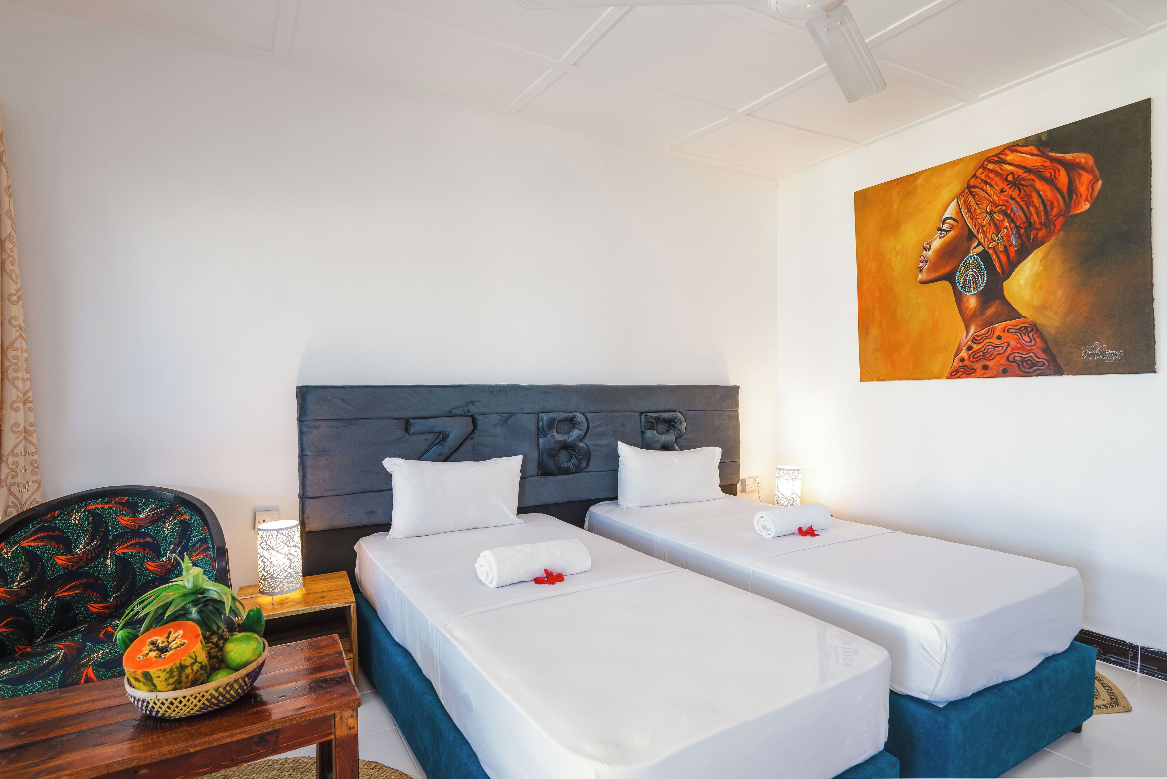 Quarto de hotel com duas camas de solteiro, uma mesinha de cabeceira de madeira, uma fruteira e um quadro na parede.