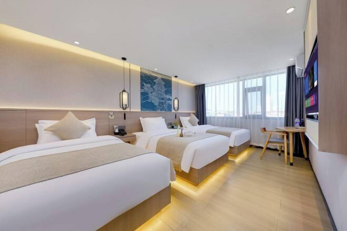 Quarto de hotel moderno com três camas, uma escrivaninha e uma janela grande.
