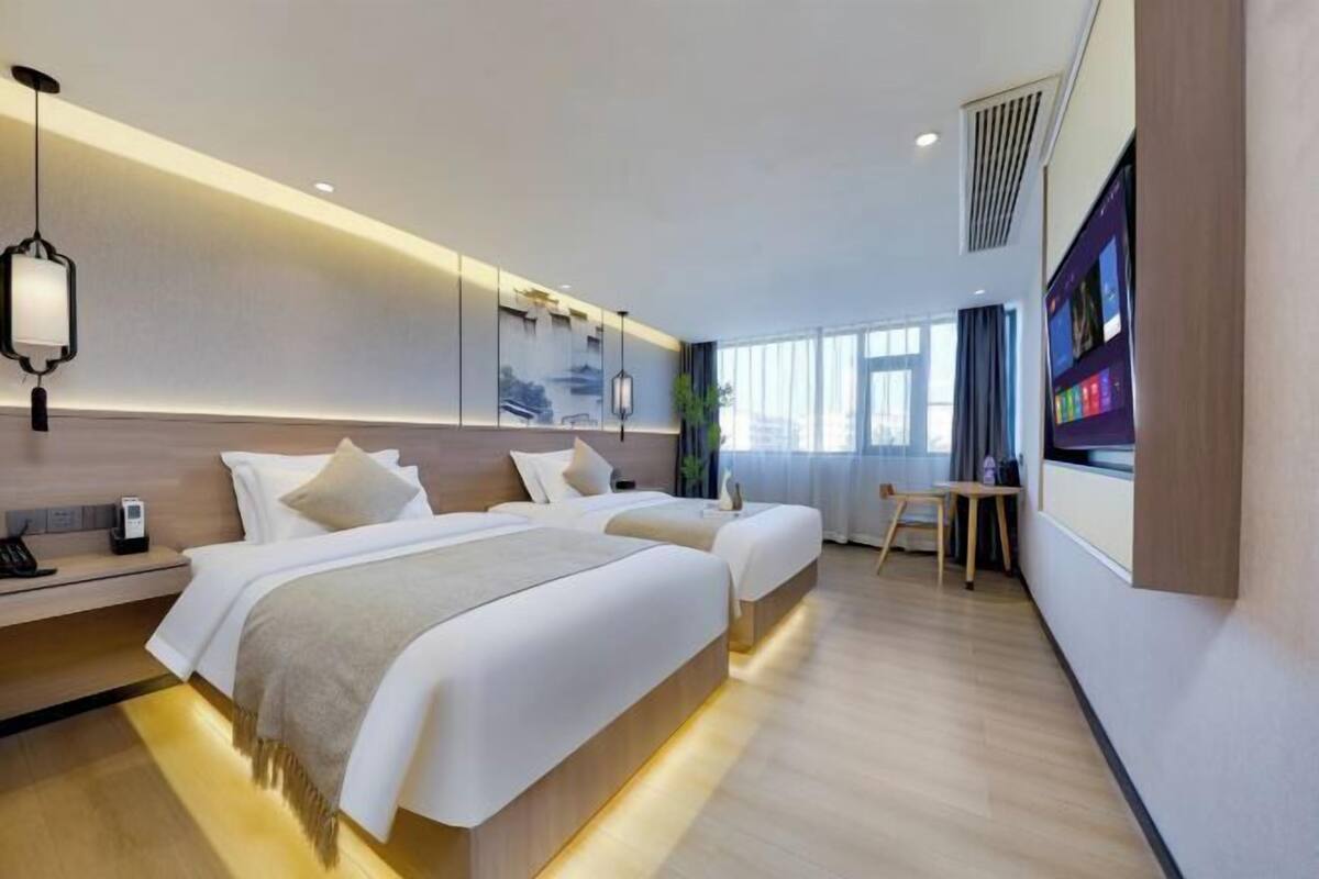 Quarto de hotel com duas camas, uma escrivaninha, uma cadeira, uma televisão e uma janela grande.