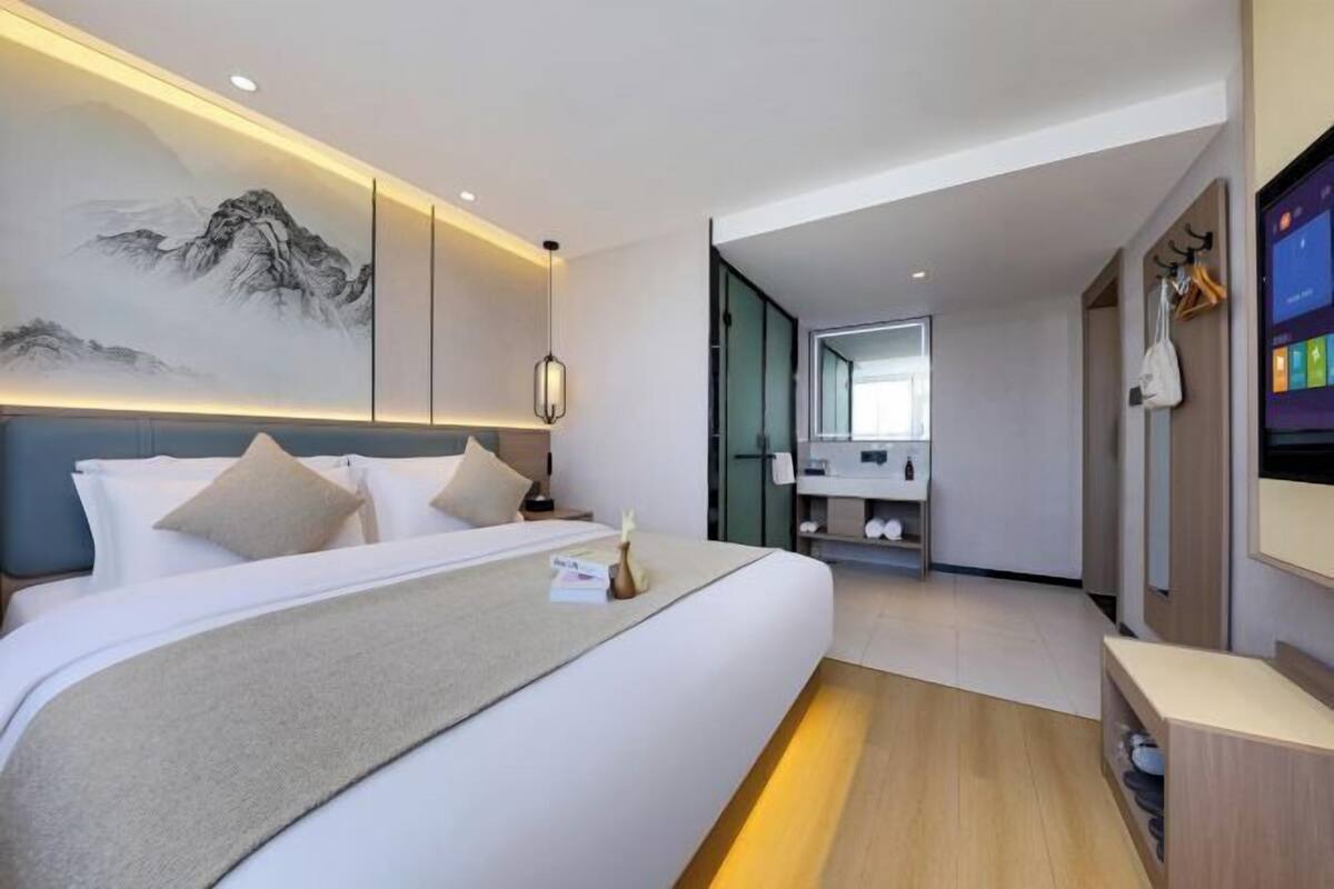 Quarto de hotel moderno com uma cama grande, uma TV fixada na parede, um cabideiro e um banheiro visível através de uma porta aberta.