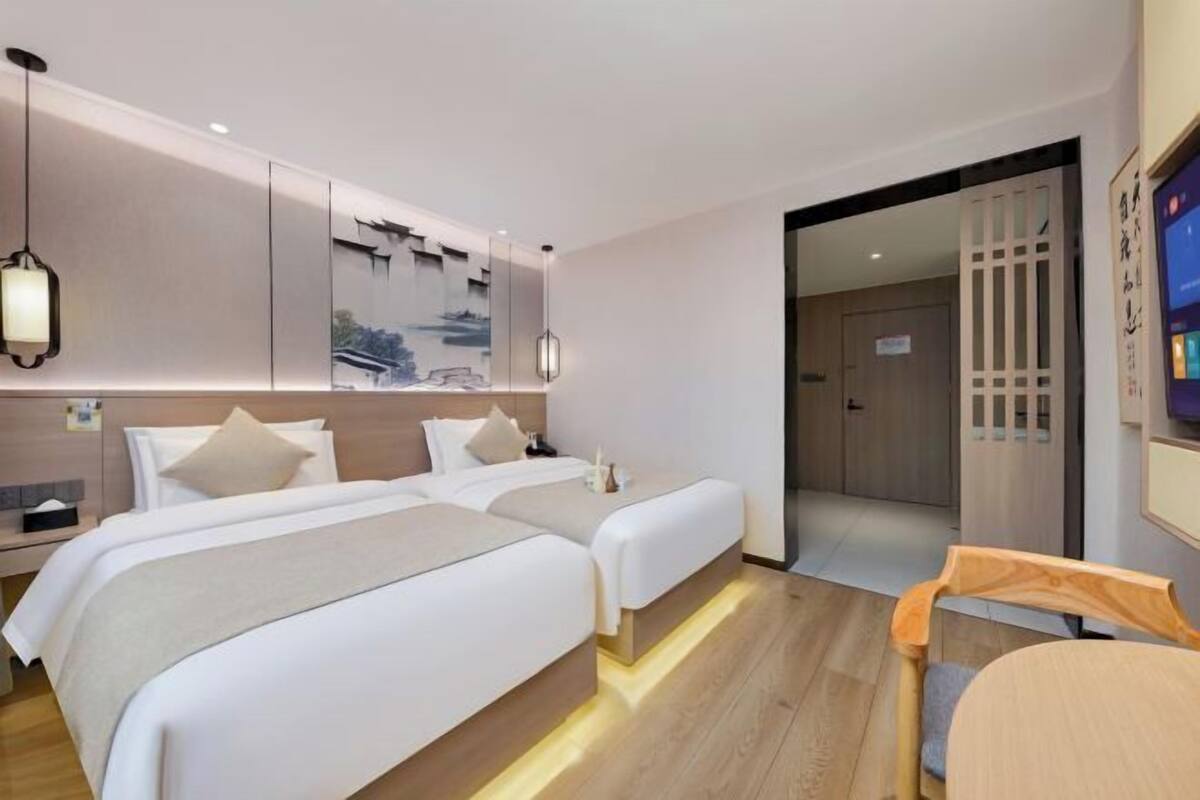 Quarto de hotel com duas camas, piso de madeira, escrivaninha com cadeira e televisão fixada na parede.