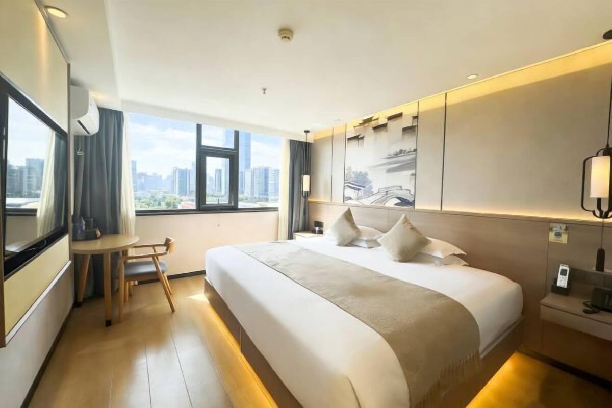 Quarto de hotel moderno com uma cama grande, uma mesa pequena, uma cadeira, uma mesinha de cabeceira e vista para a paisagem urbana através de grandes janelas.