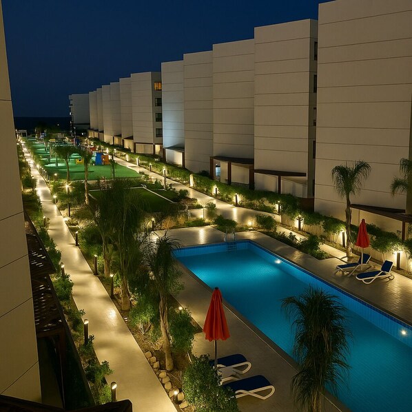 Vista noturna de uma área de piscina de hotel, com piscina, espreguiçadeiras e palmeiras.