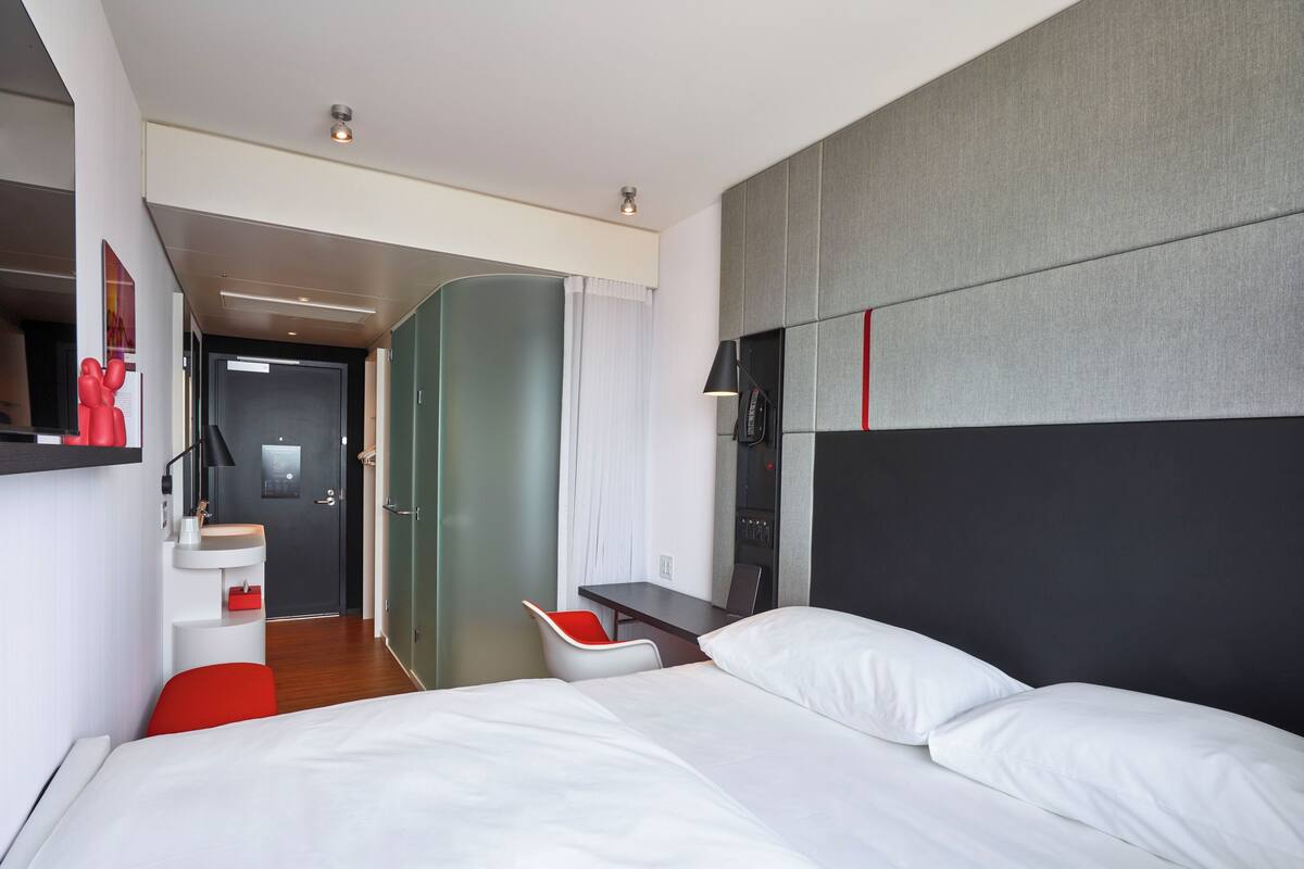 Quarto de hotel moderno com uma cama grande, uma escrivaninha e uma cadeira. É visível um banheiro com espelho e sanita através de uma porta aberta.