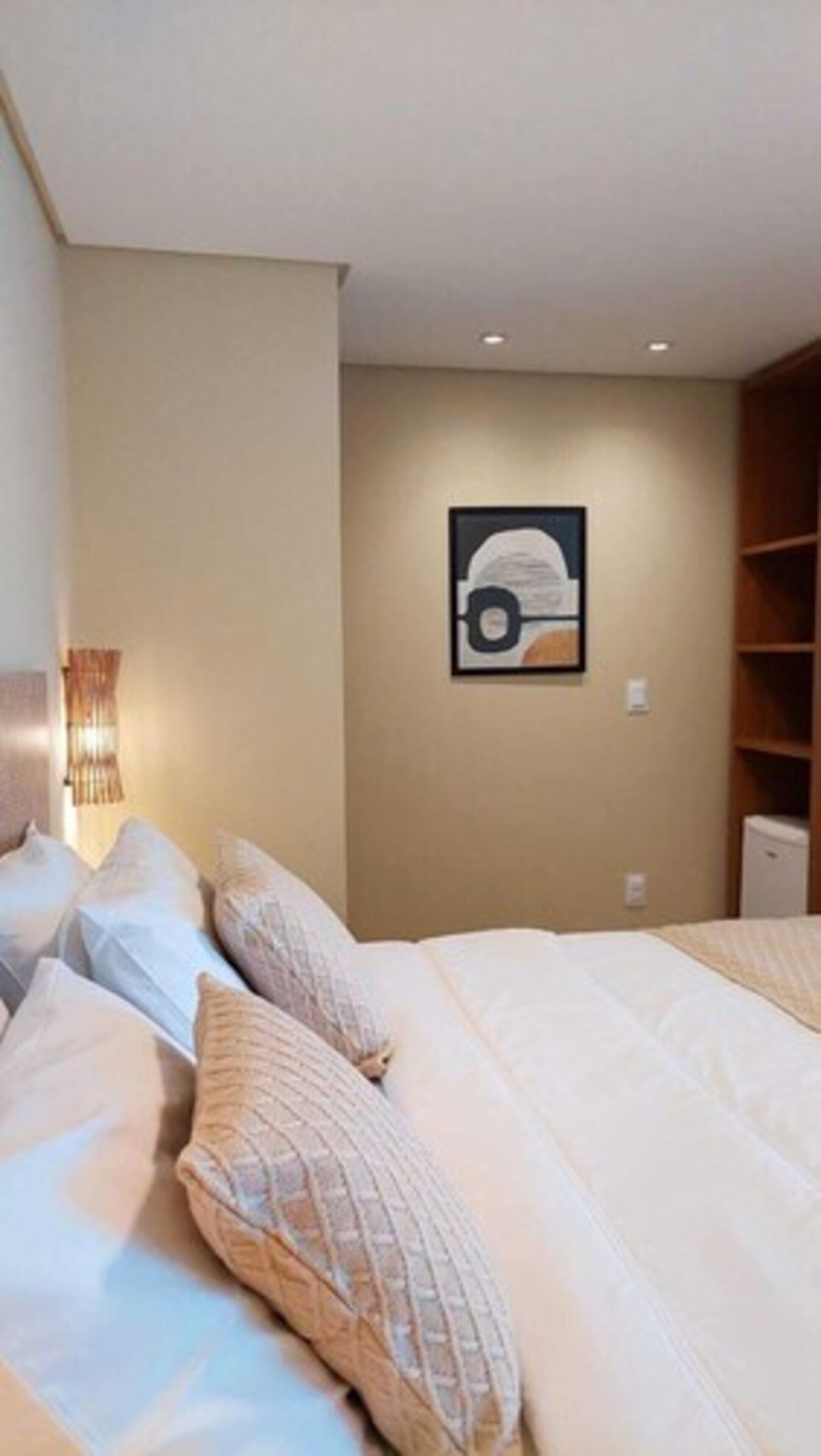 Quarto de hotel com uma cama grande, cabeceira, mesa de cabeceira, luminária e um quadro pendurado na parede.