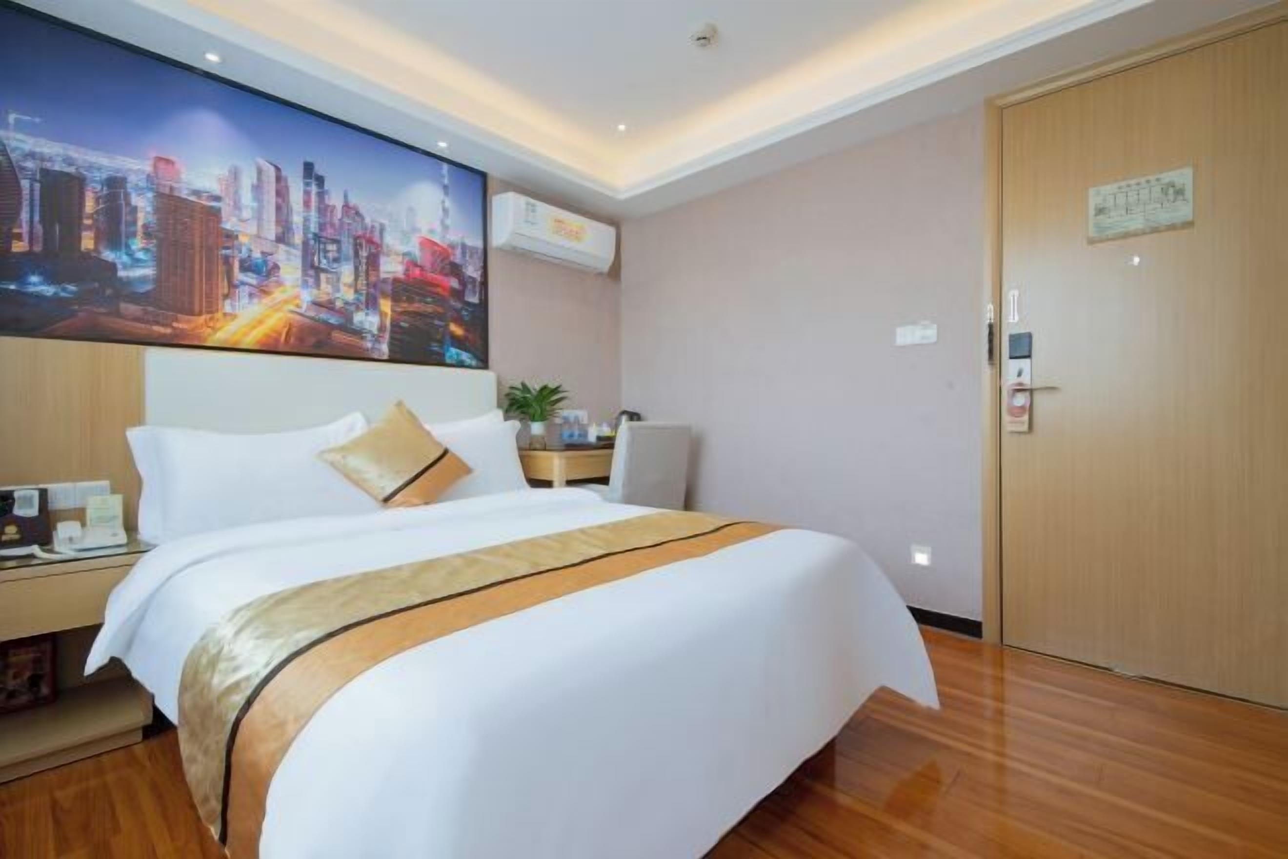 Quarto de hotel com cama, mesa de cabeceira, escrivaninha, cadeira, janela grande e vista para a cidade.