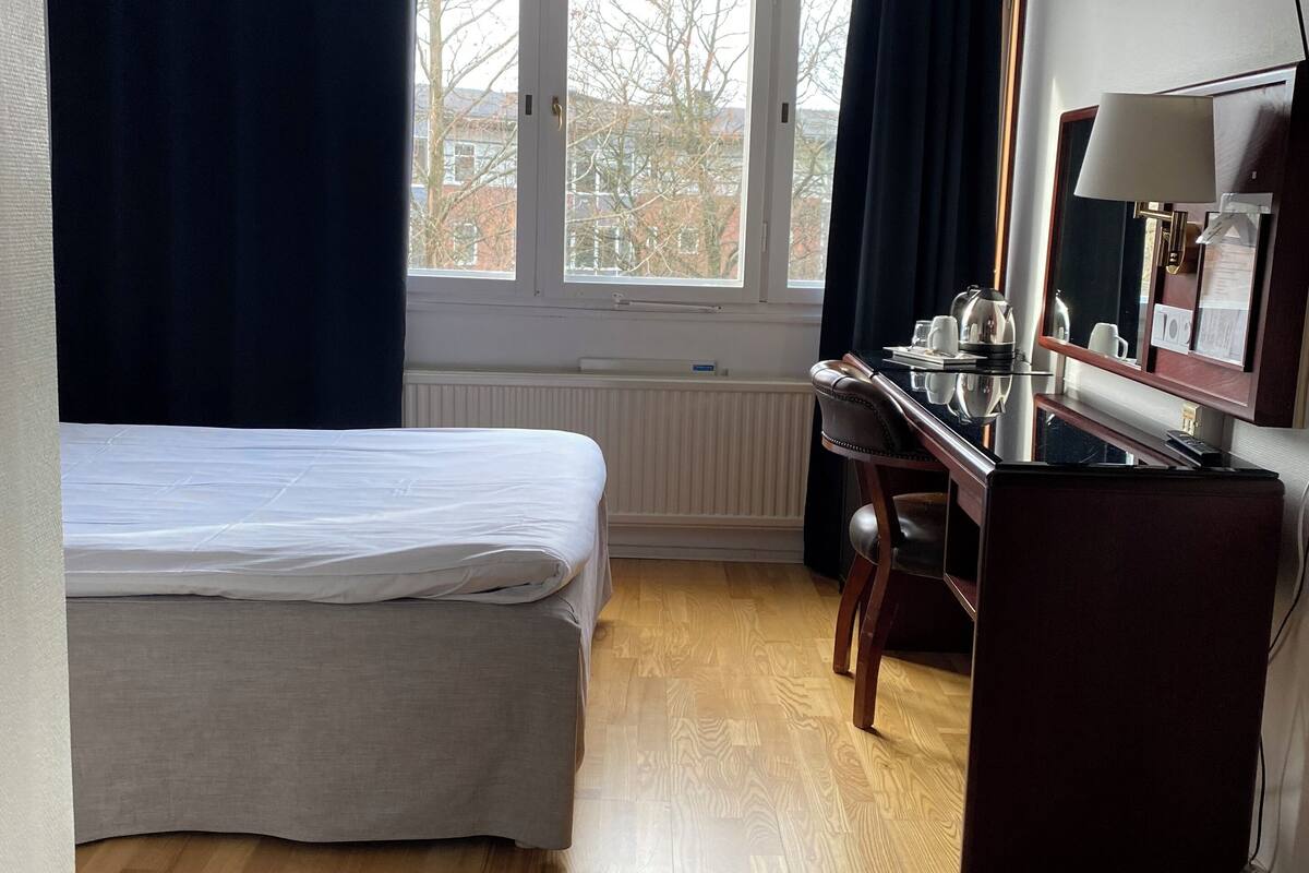 Quarto de hotel com uma cama, uma escrivaninha com cadeira, uma luminária e uma janela com cortinas.