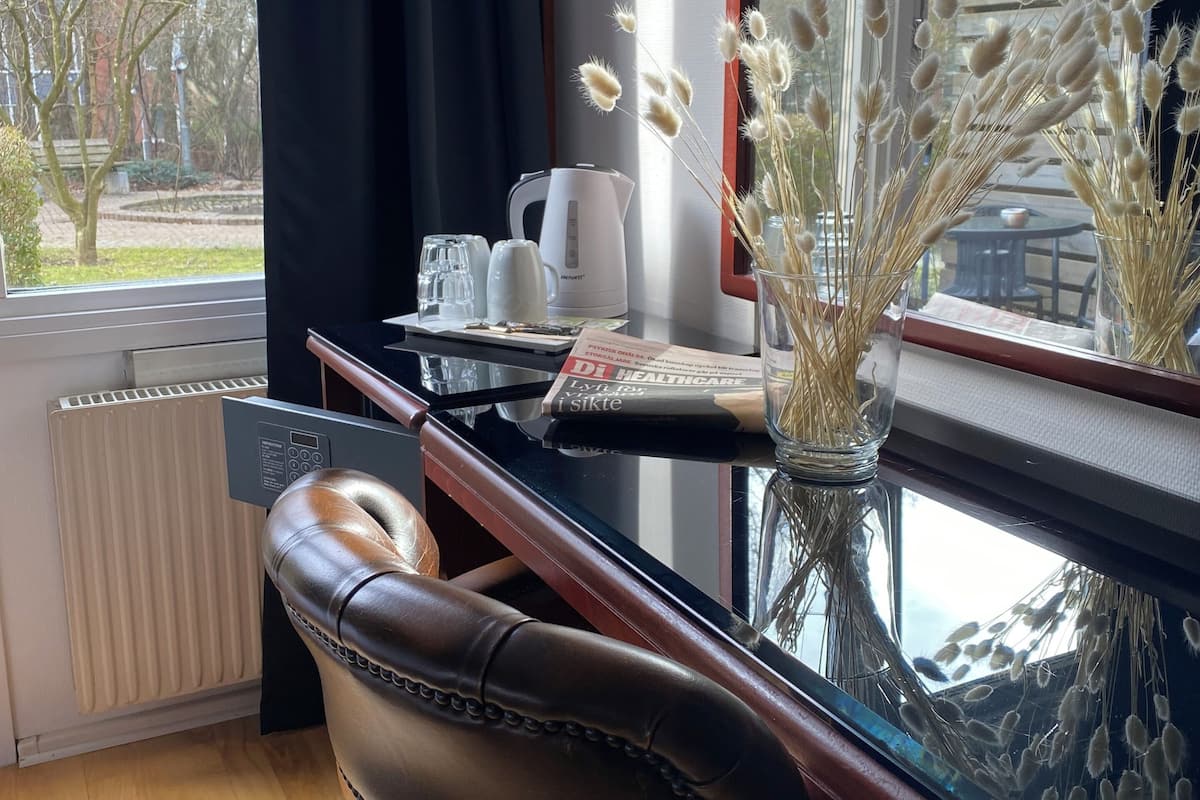 Quarto com piso de madeira, uma poltrona de couro preto, uma mesa de vidro com um vaso de flores secas, janela com vista para árvores e um edifício, e um radiador.