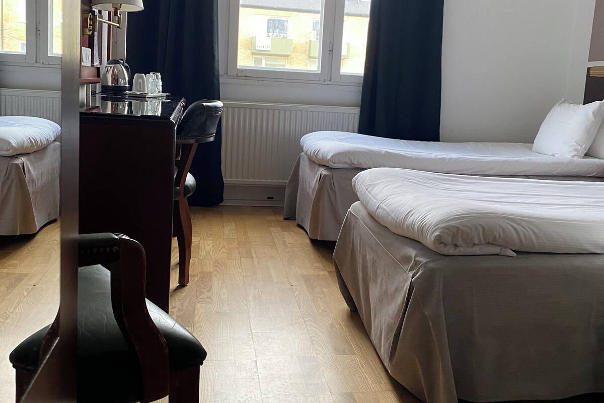 Quarto de hotel com duas camas, uma escrivaninha com abajur e vista para o exterior através de uma janela.