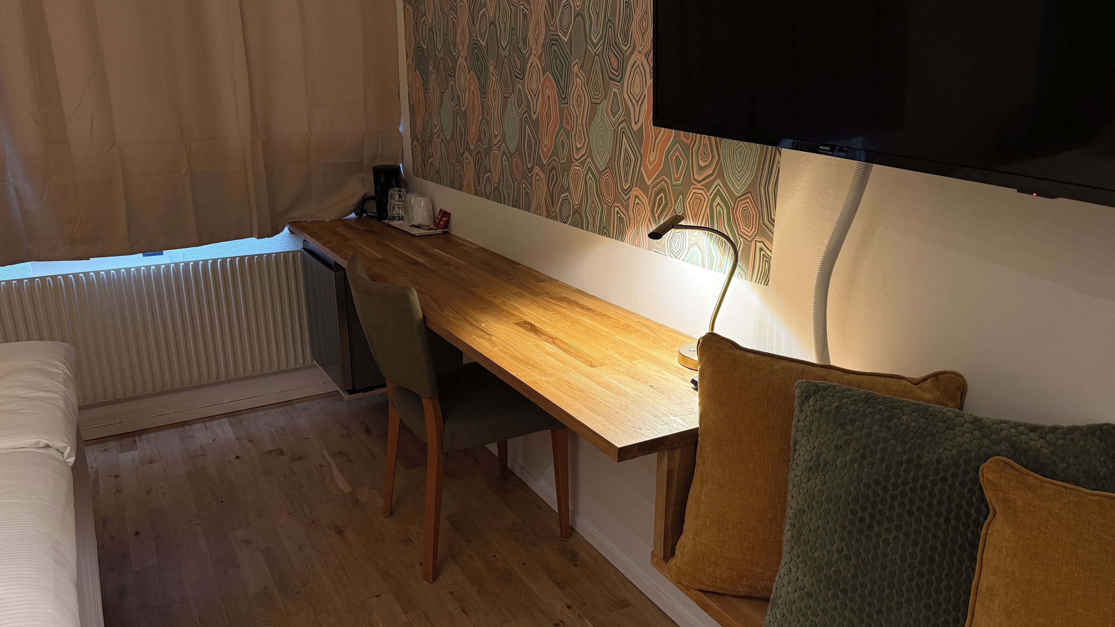 Quarto pequeno e aconchegante com escrivaninha de madeira, uma cadeira, radiador, televisão e papel de parede estampado.