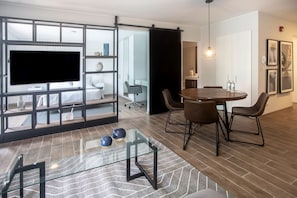 Uma sala de estar moderna com uma grande televisão de tela plana, uma mesa de centro de vidro e uma área de jantar com mesa redonda e cadeiras.