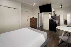 Quarto com cama, escrivaninha, cadeira, televisão e pequeno espaço de cozinha.