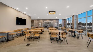 Uma ampla sala de jantar com várias mesas e cadeiras, uma TV de tela plana na parede, e grandes janelas com vista para o exterior.