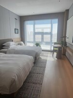 Quarto de hotel moderno com duas camas, uma janela grande, uma mesinha e um vaso de planta.