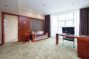 Quarto de hotel com sofá, televisão, escrivaninha e uma janela grande com cortinas.