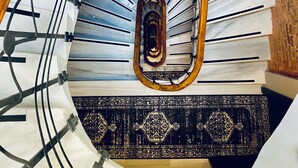 Uma escadaria em espiral com corrimão de madeira e balaústres pretos, que leva a um patamar com tapete estampado em preto e dourado.